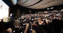 Candid photo of a conference setting, where one speaker is standing onstage with a presentation displayed behind him on a large screen, in front of an audience of several hundred people.
